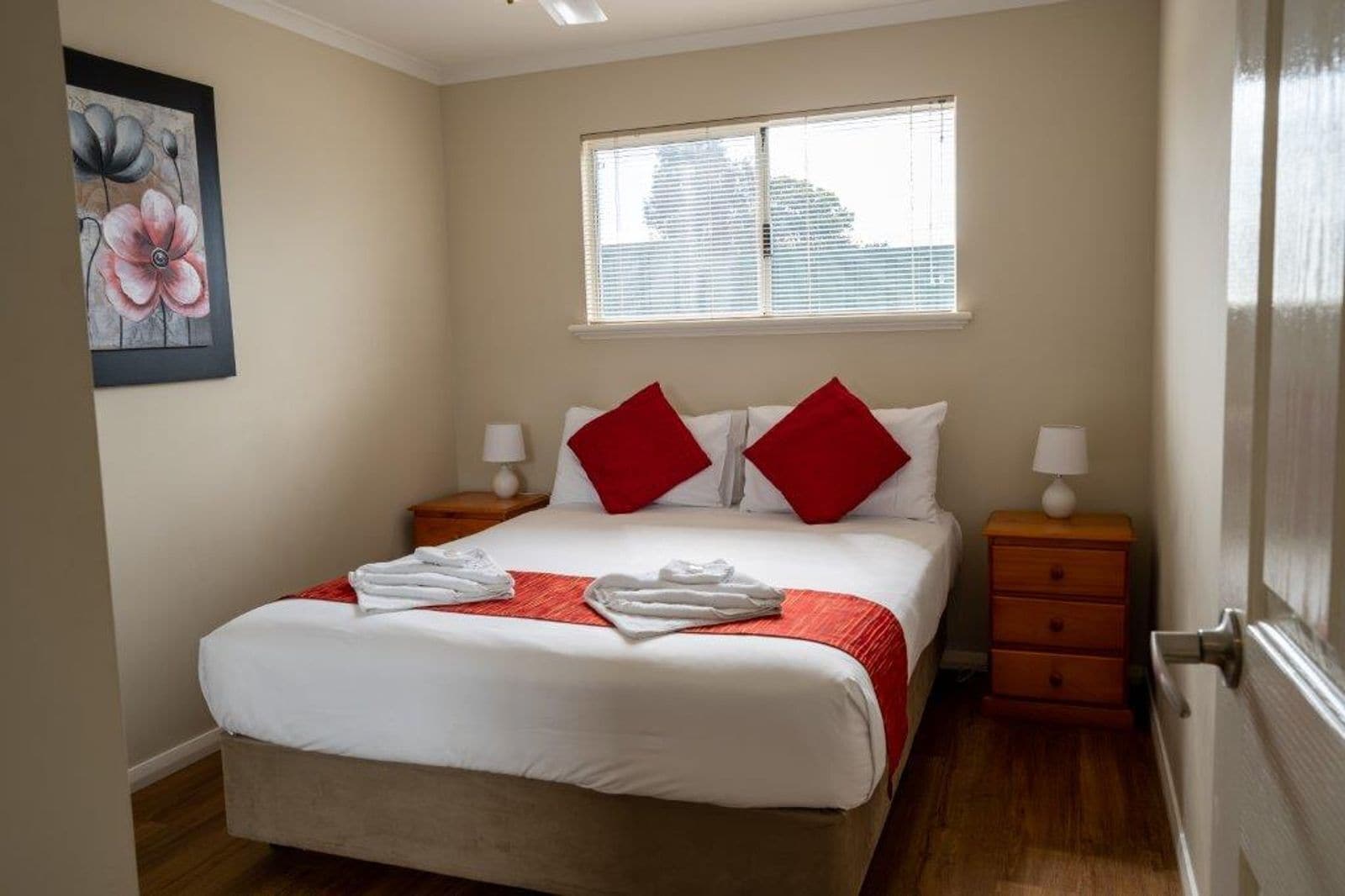 Cama bem arrumada com roupa de cama branca e vermelha, duas mesas de cabeceira com luminárias e uma janela com persianas.