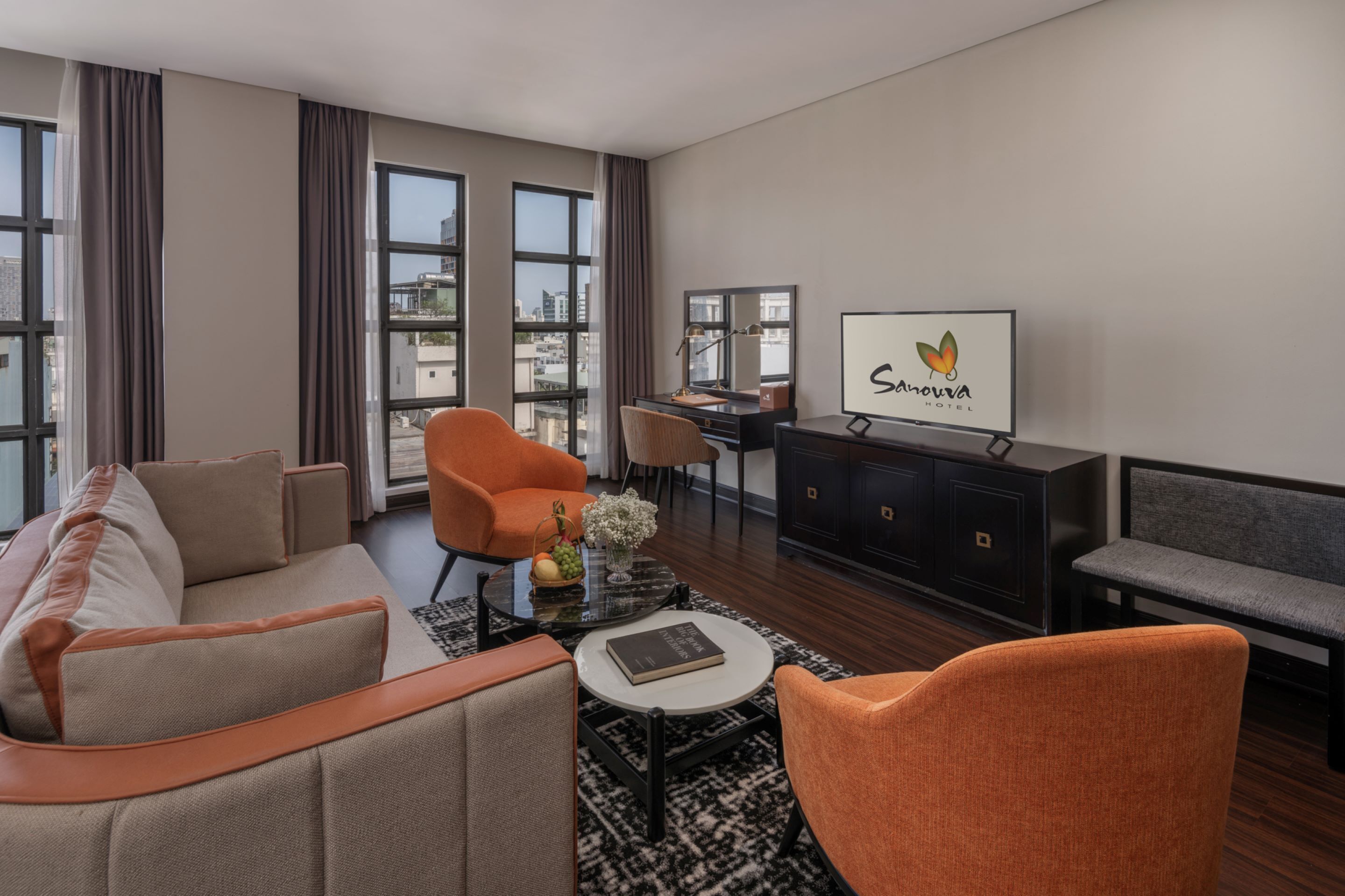 Quarto de hotel moderno com sofá, poltronas, escrivaninha e televisão exibindo o nome do hotel.