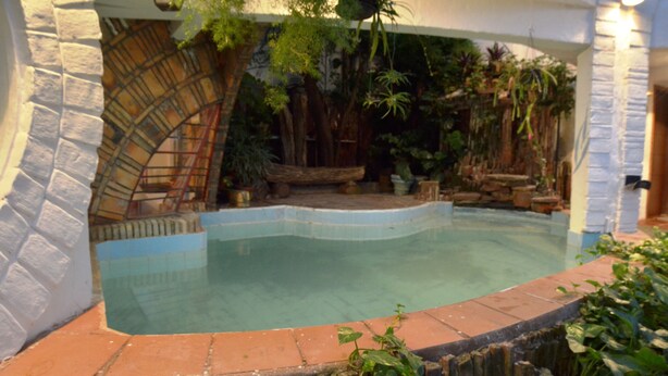 Piscina com muro de pedra e arco de madeira, rodeada por vegetação exuberante e plantas.