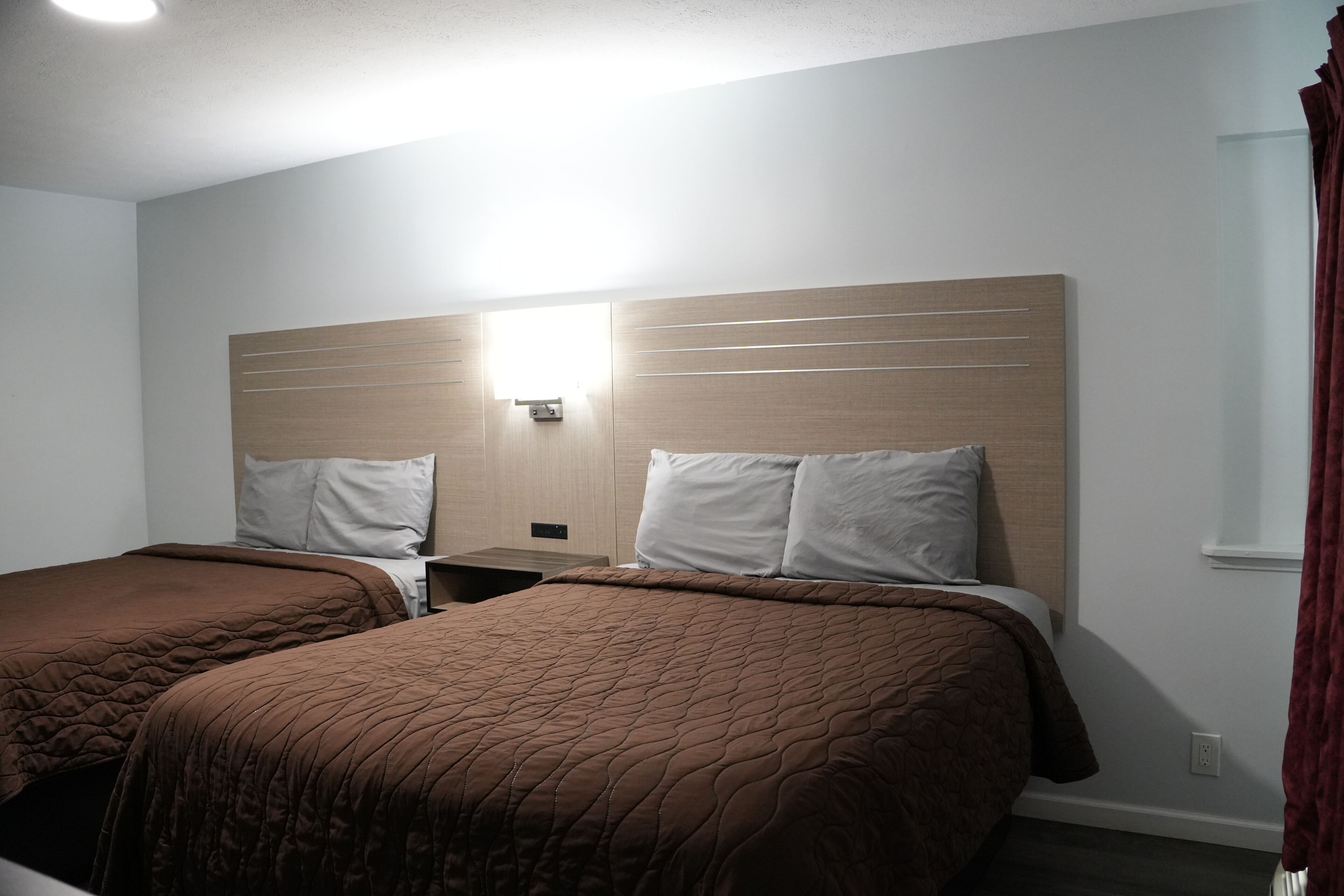 Quarto de hotel com duas camas, uma mesa de cabeceira, uma luminária na parede e uma janela com cortinas.