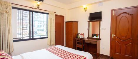 Quarto de hotel com cama, escrivaninha, guarda-roupa, televisão e janela com cortinas.