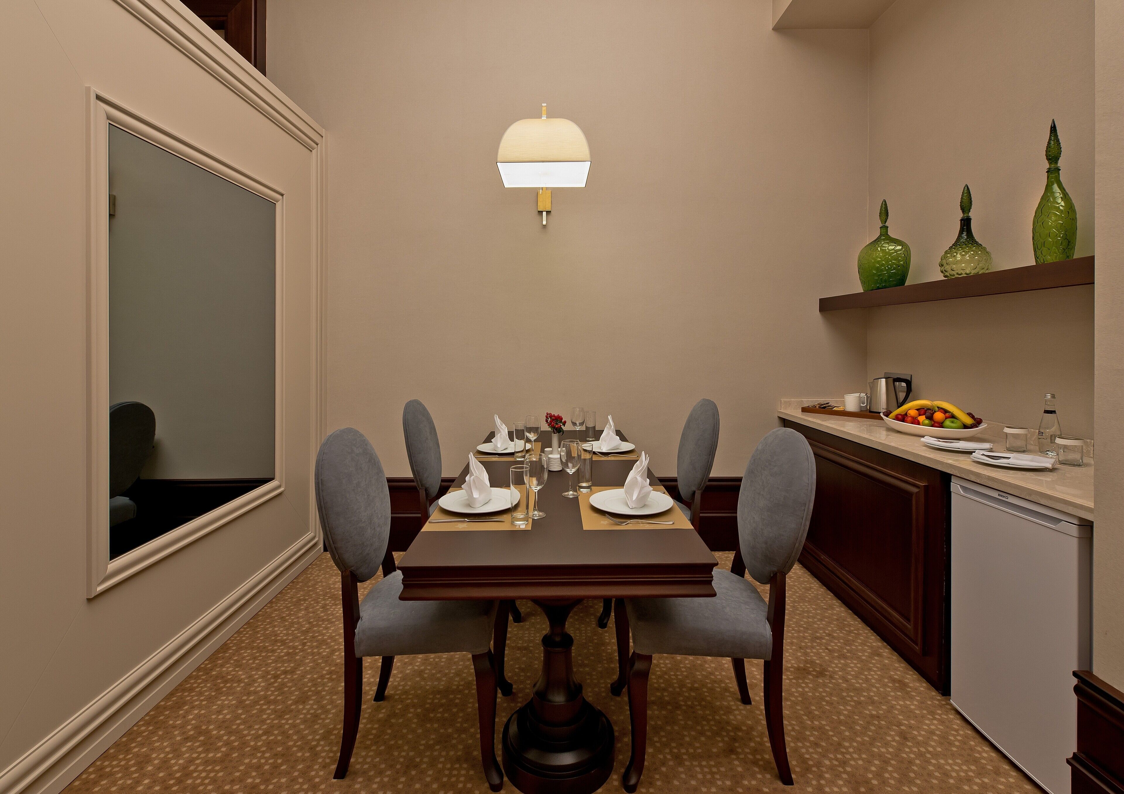 Uma sala de jantar com uma mesa posta para quatro pessoas, cadeiras, luminária fixada na parede e uma tigela de frutas em uma bancada.