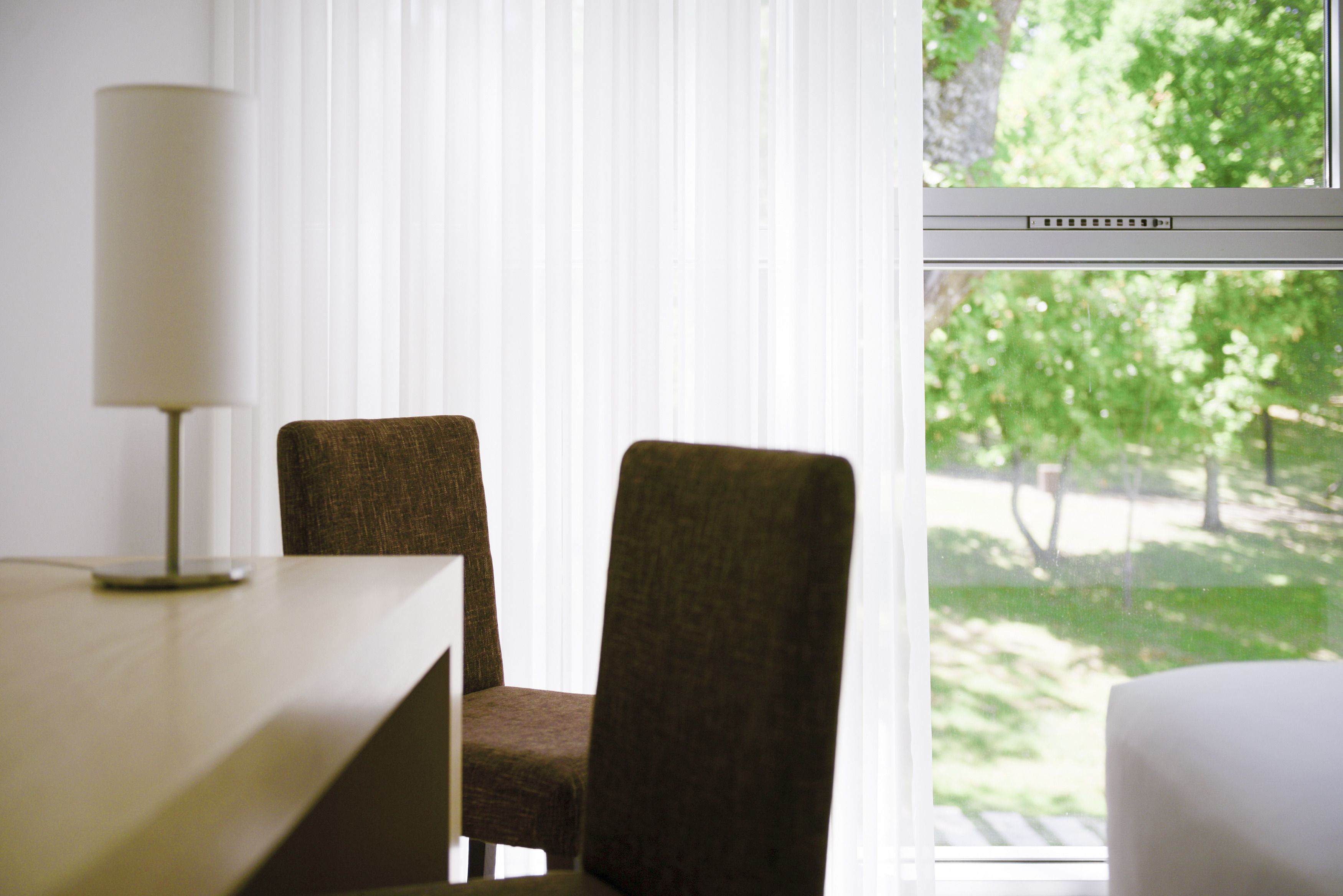 Quarto com mesa, duas cadeiras, abajur e janela com vista para vegetação.