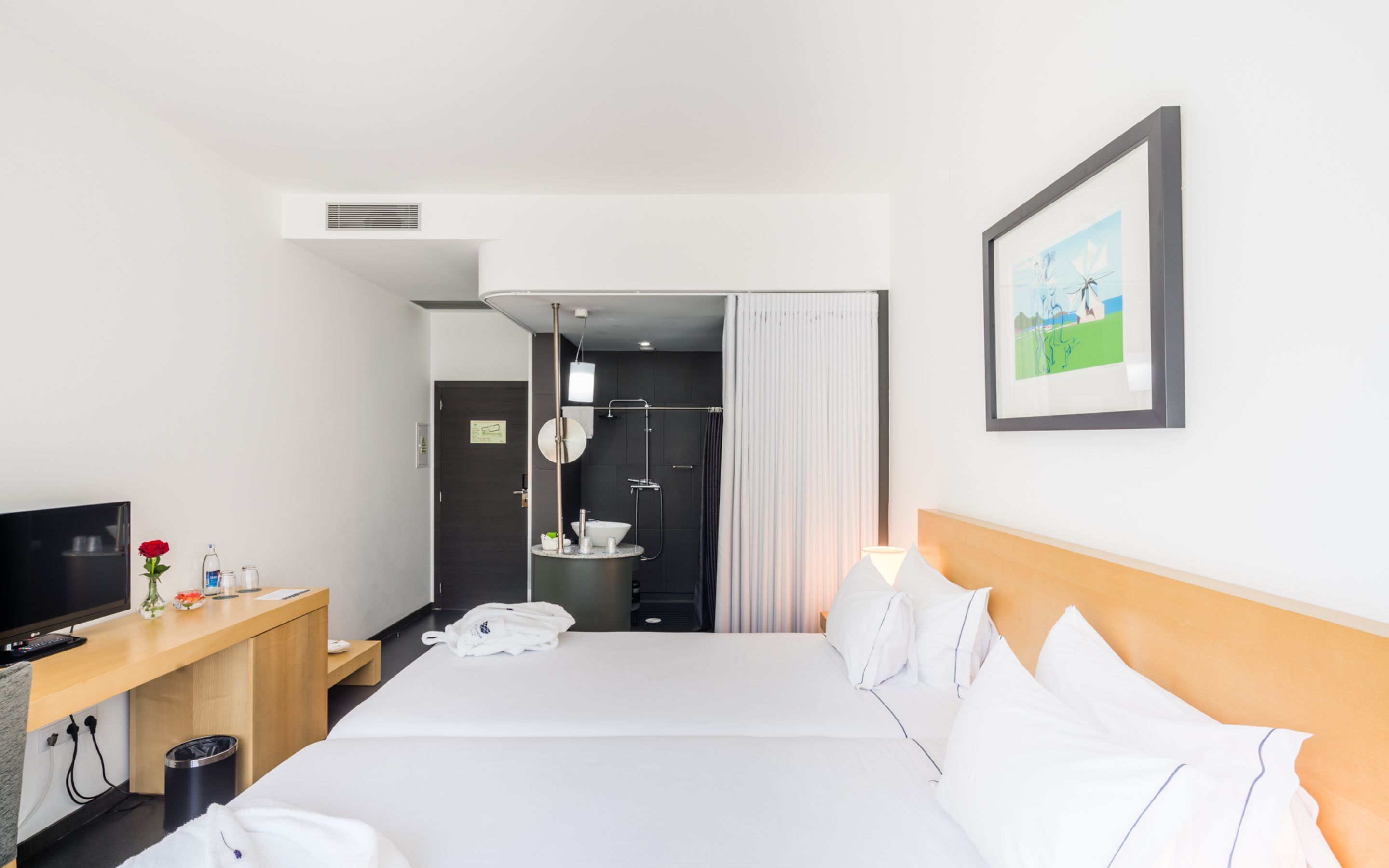 Quarto de hotel com cama, secretária com televisão, chuveiro e quadro na parede.