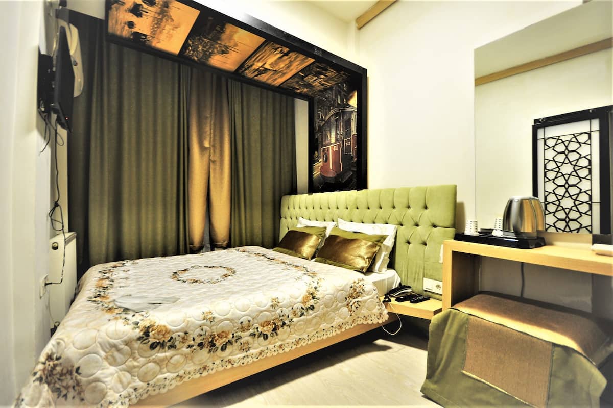 Um quarto com cabeceira estofada, cama com colcha estampada floralmente, um banquinho estofado verde e uma mesa de cabeceira com abajur.