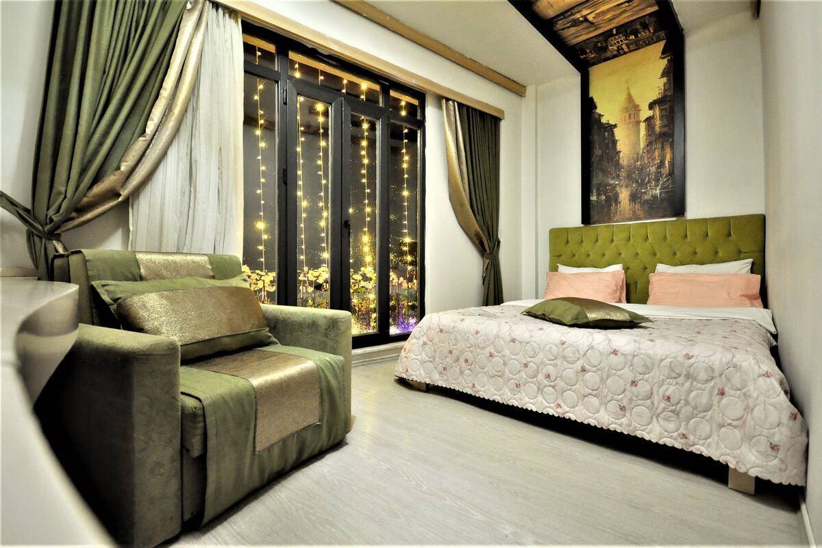 Um quarto com uma poltrona estofada verde, uma cama com colcha branca e uma janela grande com cortinas.