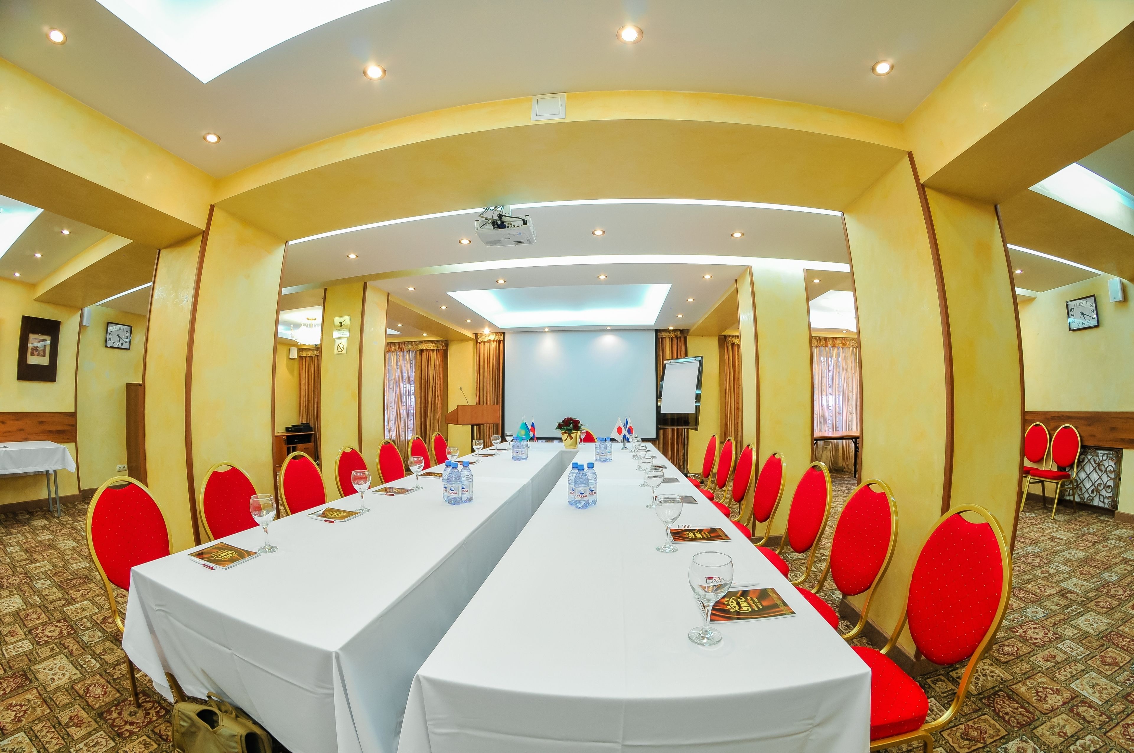 Uma sala de conferências com uma mesa comprida, cadeiras vermelhas e uma tela de projetor.