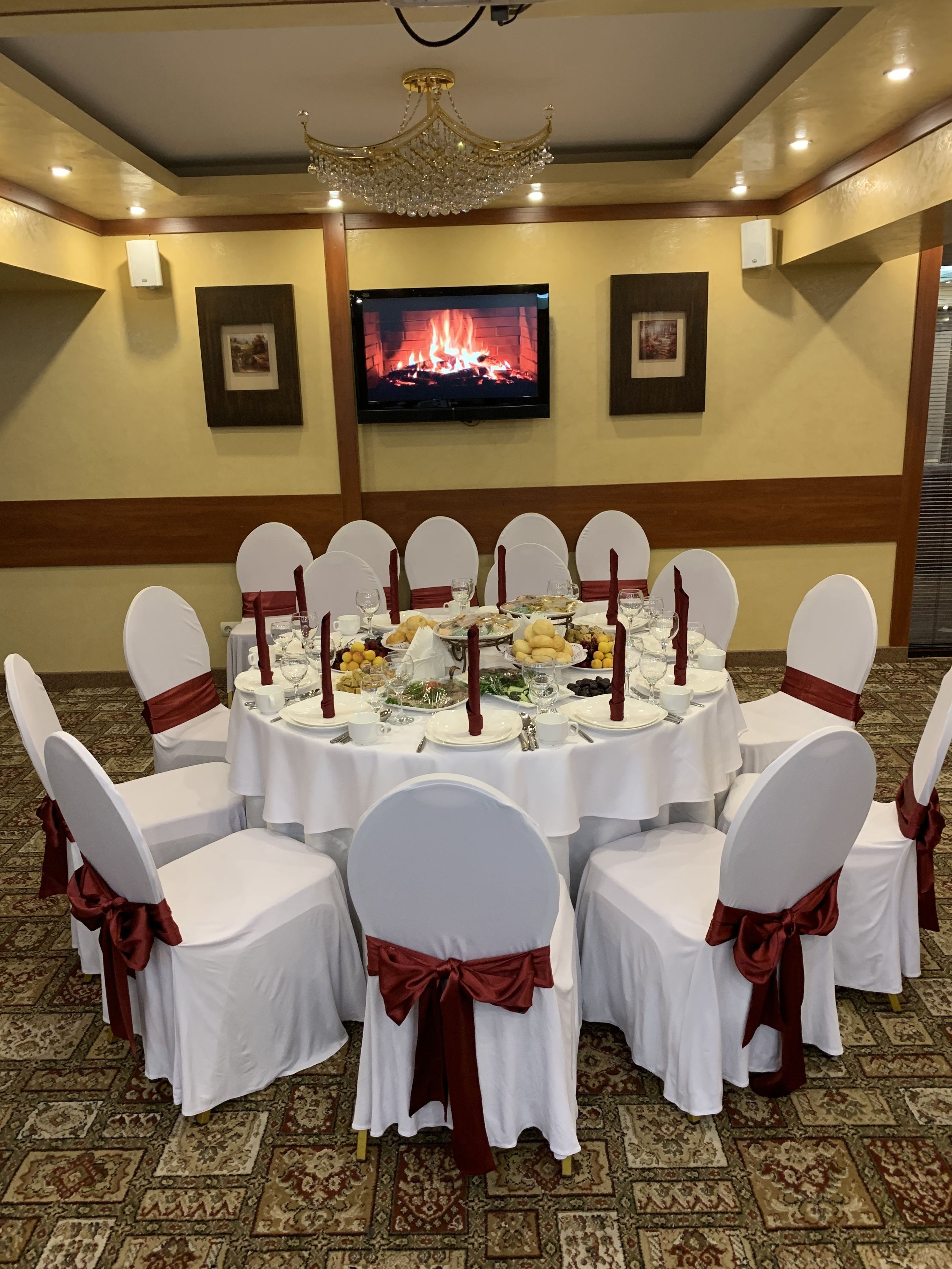 Uma sala de banquetes com mesas redondas, cadeiras brancas com fitas vermelhas, um candelabro e uma lareira na parede.