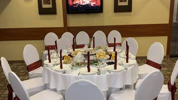 Uma sala de banquetes com mesas redondas, cadeiras brancas com fitas vermelhas, um candelabro e uma lareira na parede.