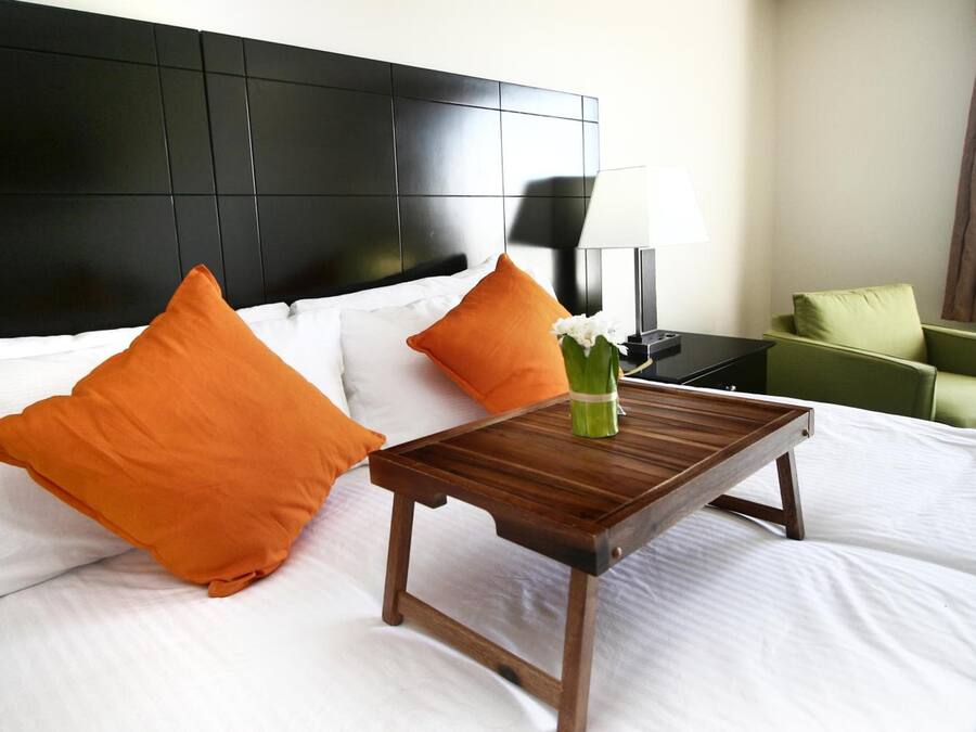 Quarto de hotel com uma cama grande, duas almofadas laranja, uma mesa de cabeceira de madeira, uma cadeira verde e um abajur.