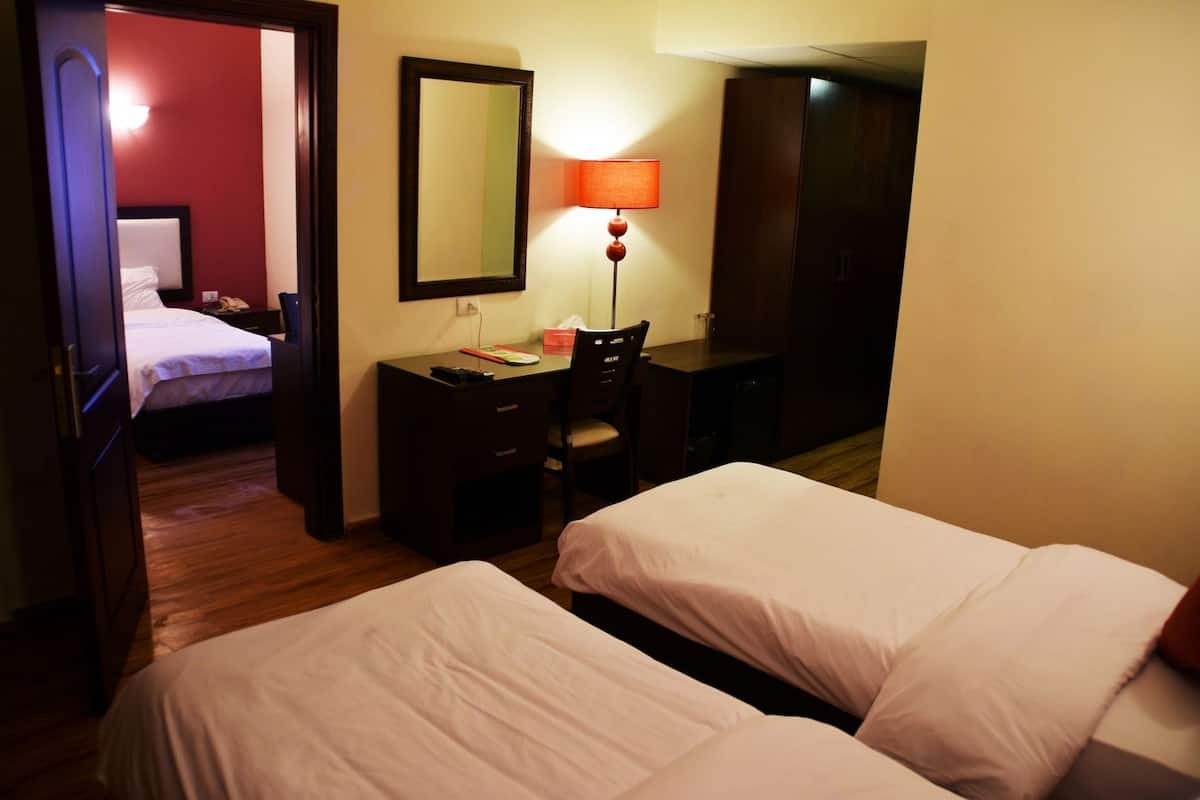 Quarto de hotel com duas camas, uma escrivaninha com cadeira, uma luminária e um espelho.