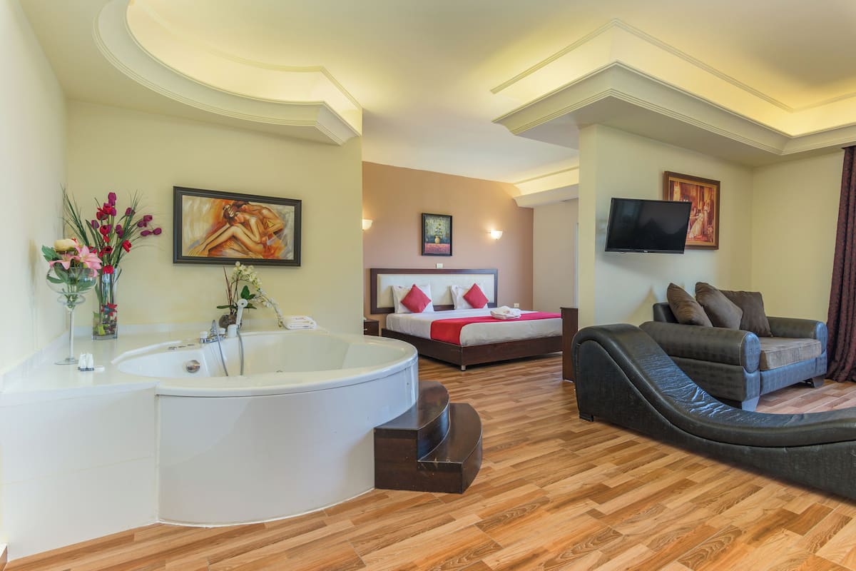 Quarto de hotel moderno com uma banheira grande, uma cama com almofadas vermelhas, um sofá e uma televisão de tela plana.