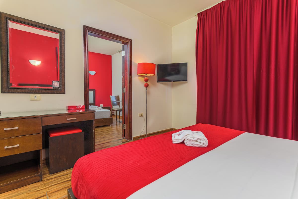 Quarto de hotel com cama, escrivaninha com gavetas, espelho, televisão e cortina vermelha.