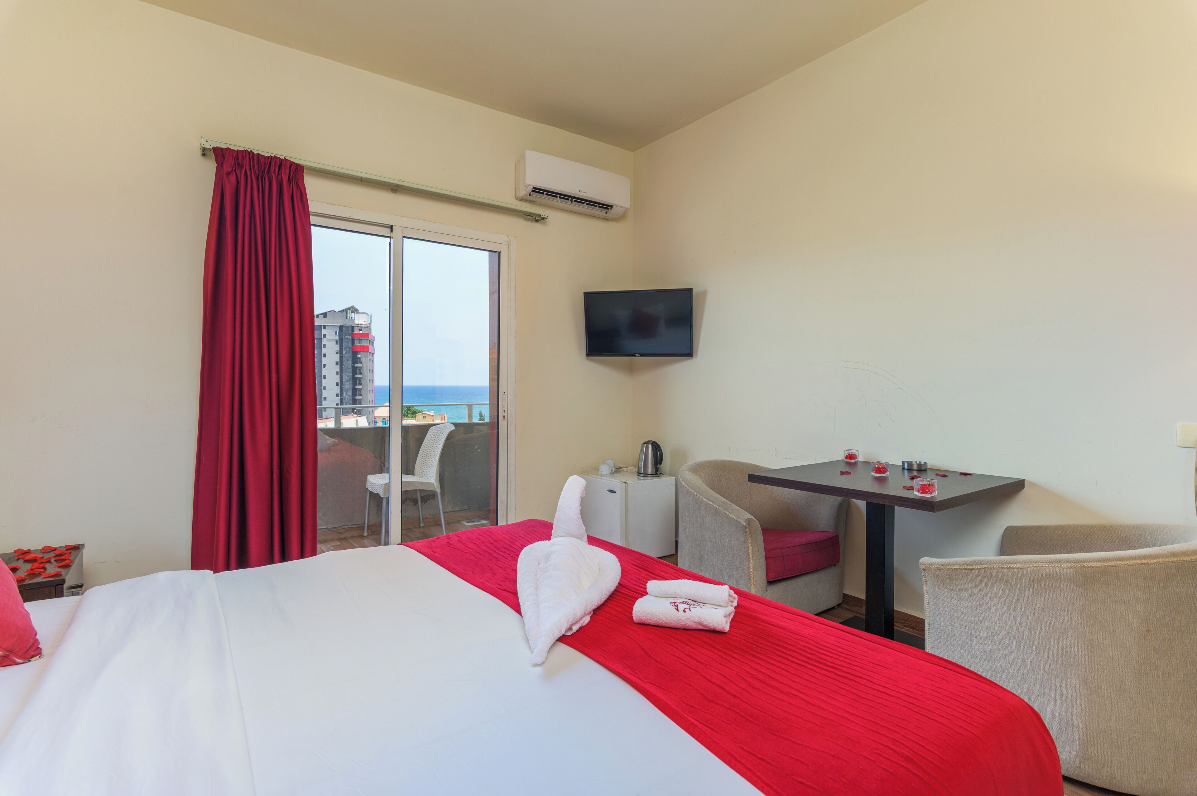 Quarto de hotel com cama, televisão, uma pequena mesa com chaleira, cadeira e varanda com cortinas vermelhas.