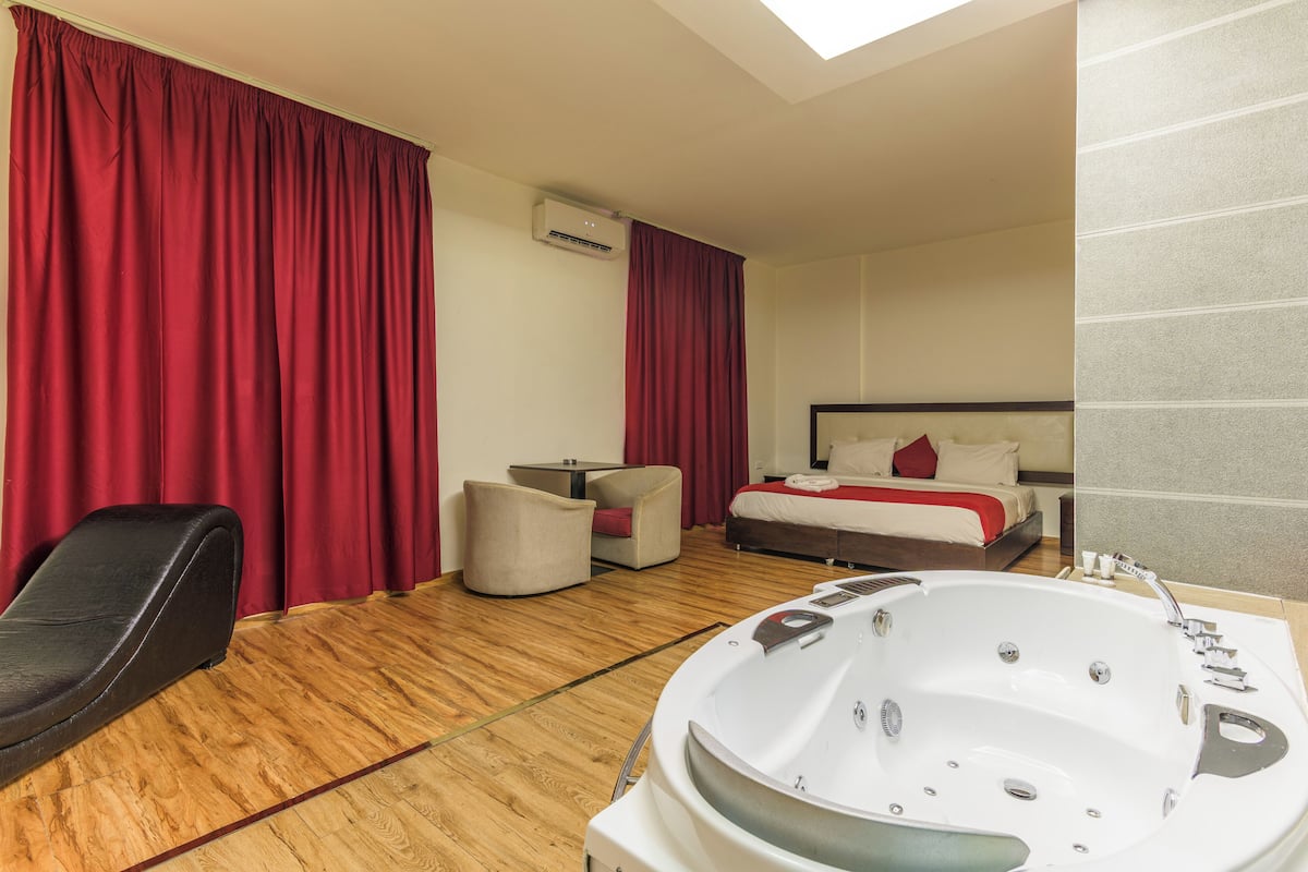Quarto de hotel com uma cama grande, um espaço de estar com uma poltrona e uma mesinha, uma banheira ampla e cortinas vermelhas.