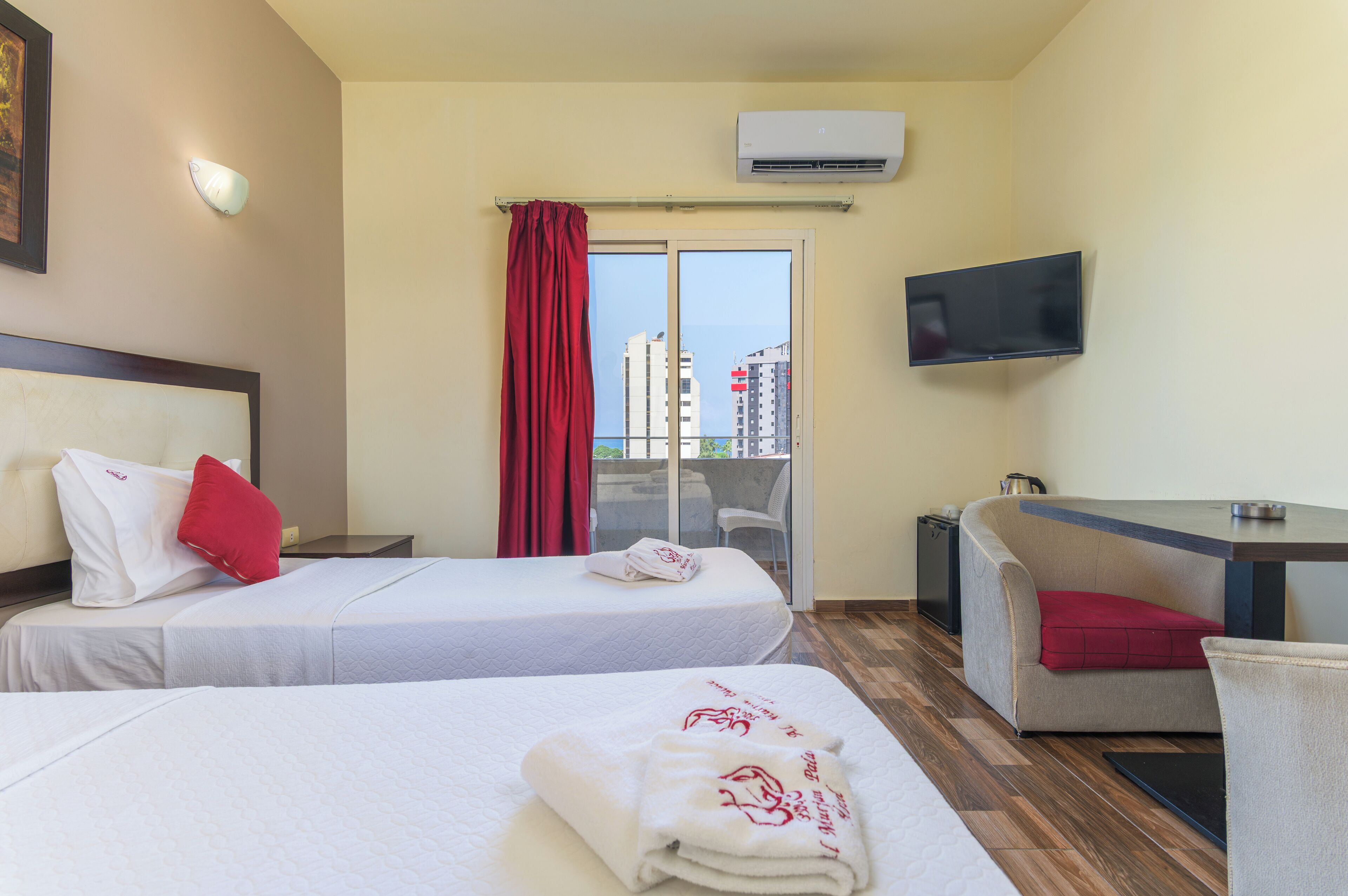 Quarto de hotel com duas camas, televisão, escrivaninha e vista para edifícios pela janela.