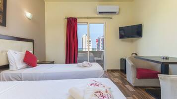 Quarto de hotel com duas camas, televisão, escrivaninha e vista para edifícios pela janela.