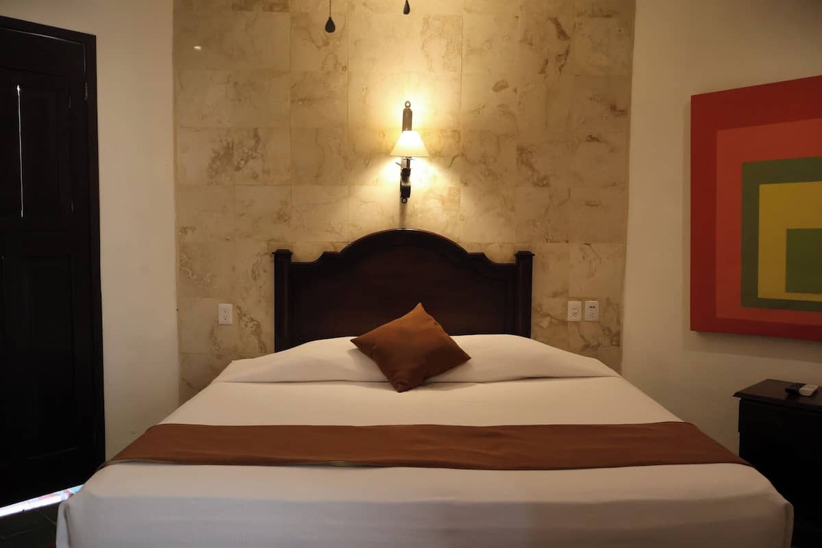 Quarto de hotel com cama, luminária na parede, tomada e obra de arte vermelha e amarela.