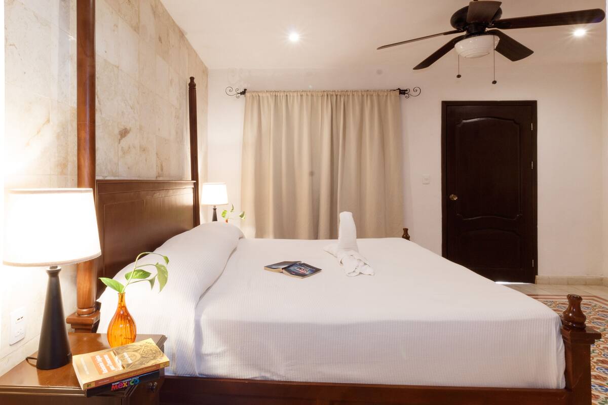 Quarto de hotel com uma cama, mesa de cabeceira com luminária, vaso com planta e uma porta.