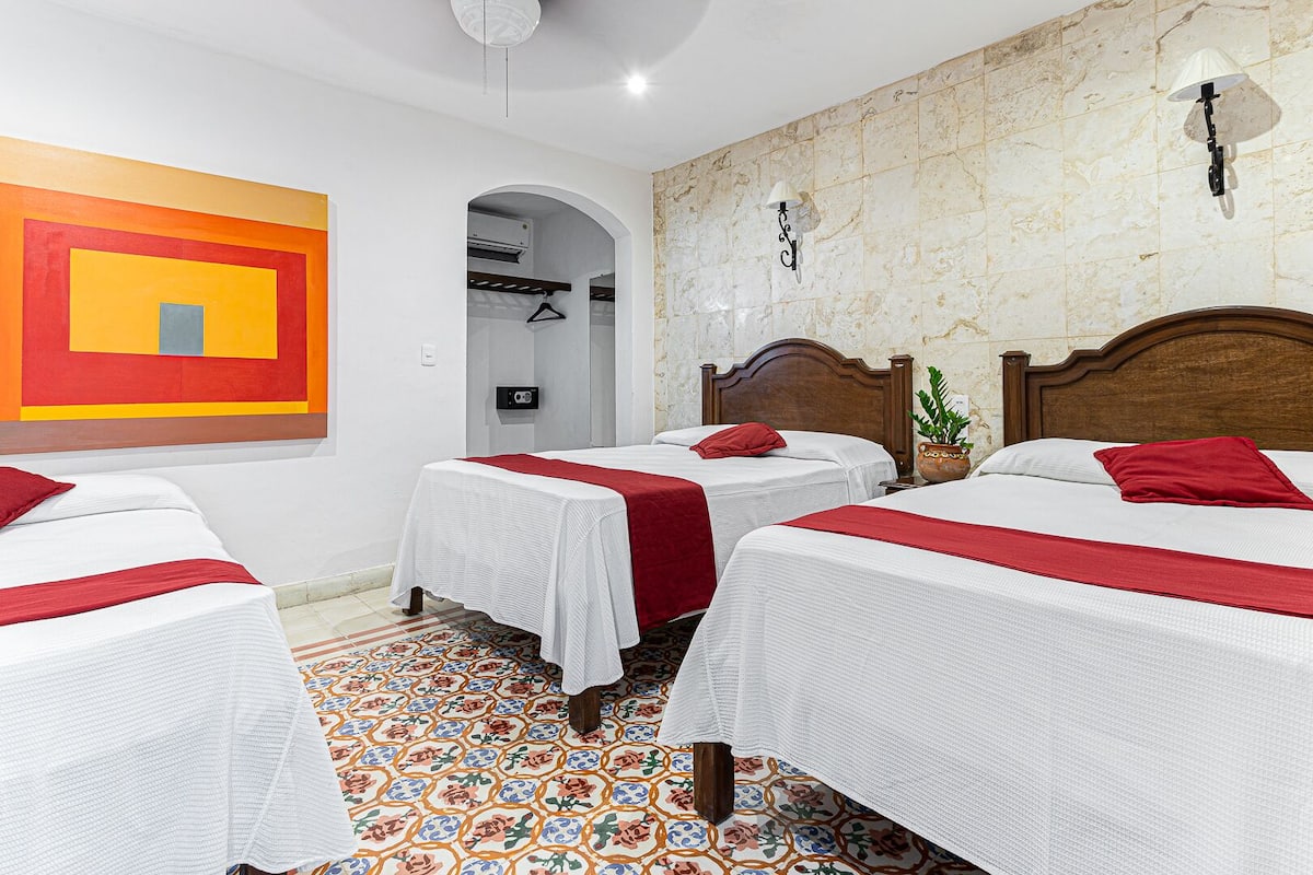 Quarto de hotel com três camas, um quadro abstrato grande, piso de azulejos e cabeceira de madeira.