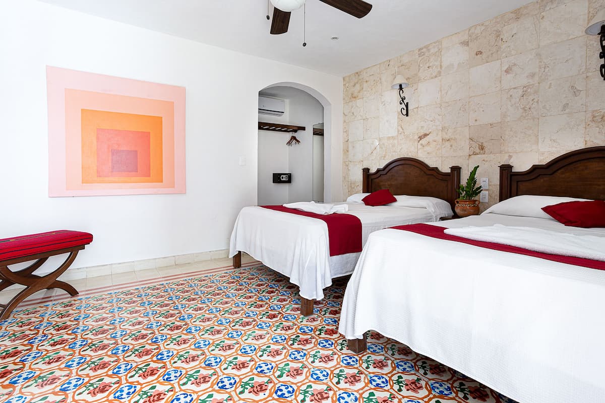Quarto de hotel com duas camas, um banco vermelho, um chão estampado e uma parede com uma pintura geométrica.