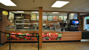 Interior de um restaurante de fast food com balcão, painéis de menu, máquina de Pepsi e uma vitrine com frutas e vegetais frescos.