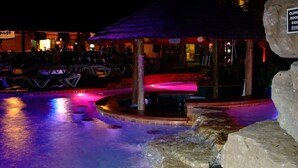 Uma área de piscina à noite com um gazebo de colmo, cadeiras de descanso e uma banheira de hidromassagem.