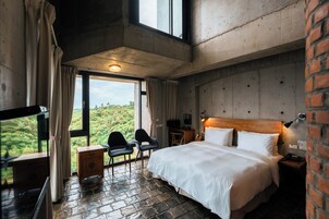 Quarto de hotel com uma cama grande, uma escrivaninha, duas cadeiras, uma mesa pequena e vista para o verde pela janela.