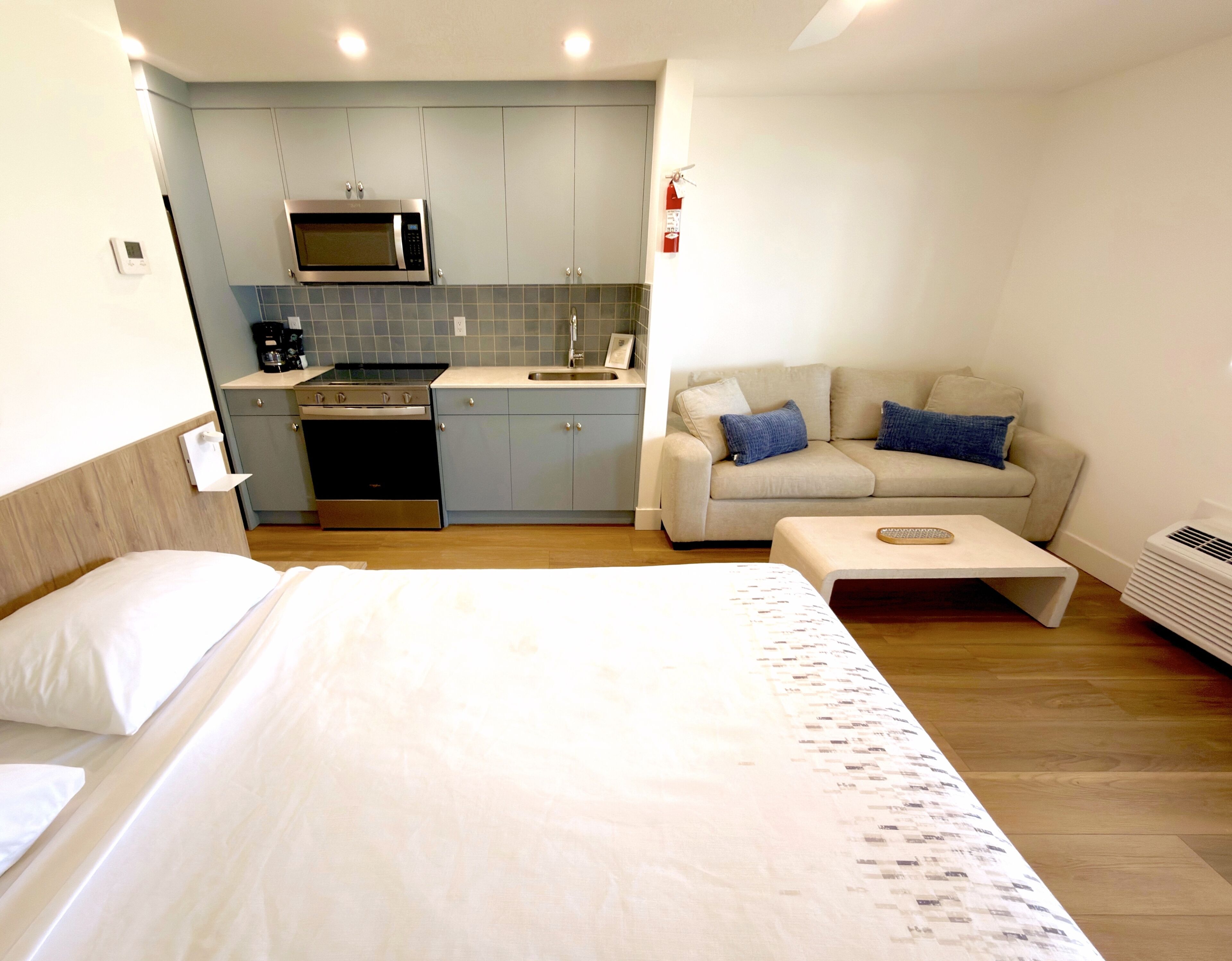 Quarto de hotel compacto com cama, sofá, pequeno espaço de cozinha e máquina de café.