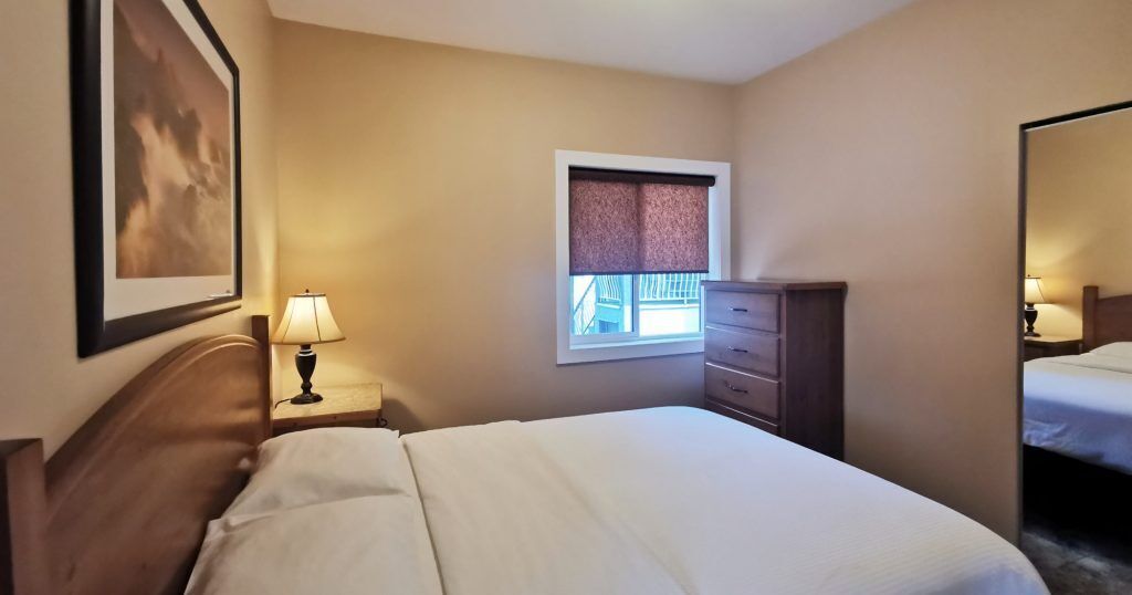 Quarto de hotel com cama, mesa de cabeceira com abajur, penteadeira, janela com persianas e um quadro na parede.