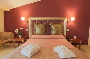 Quarto de hotel com uma cama grande, duas almofadas douradas, uma mesinha de cabeceira de madeira com um telefone e um abajur, uma poltrona floral e um vaso com flores.