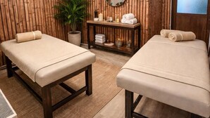 Sala de spa com duas mesas de massagem, parede de madeira, espelho, planta e luminária no teto.