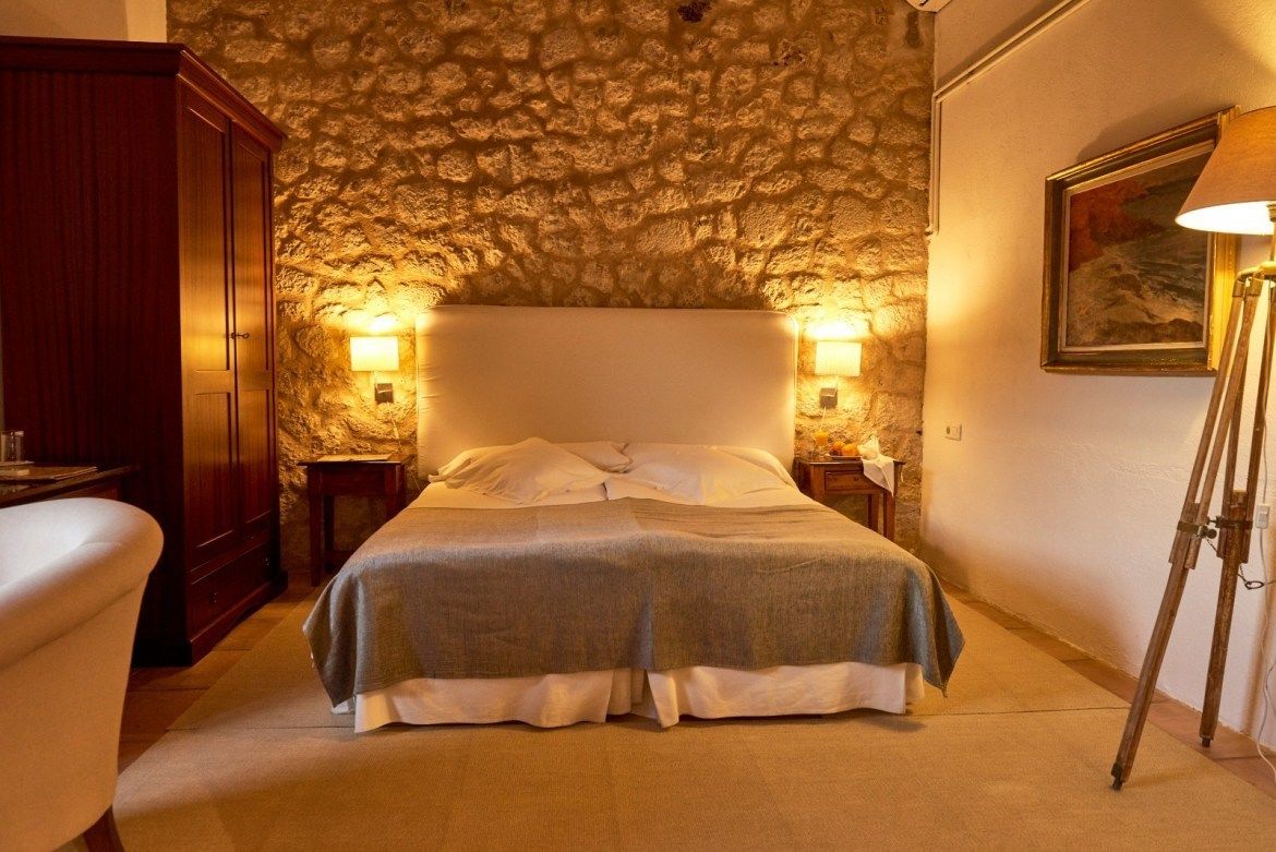 Quarto com parede de pedra, cama com cobertor cinzento, duas luminárias de apoio, guarda-roupa de madeira e um abajur.