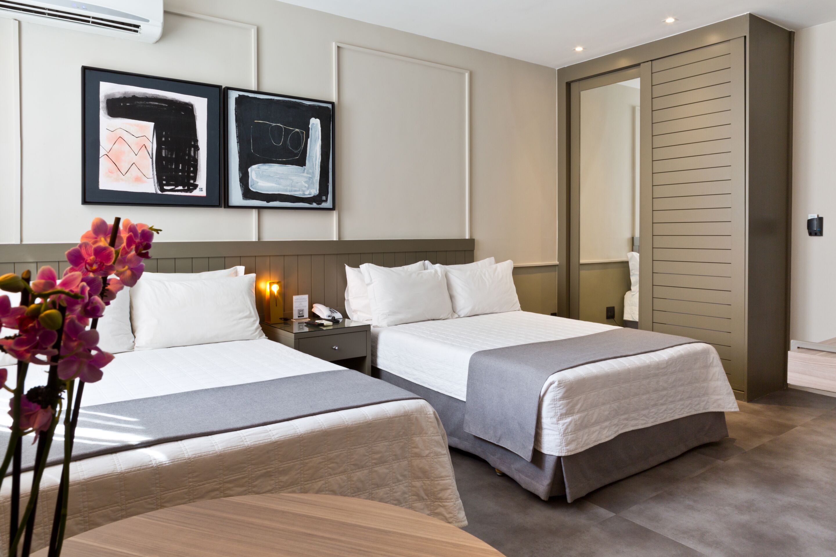 Quarto de hotel com duas camas, uma mesinha de cabeceira, um espelho e obras de arte na parede.