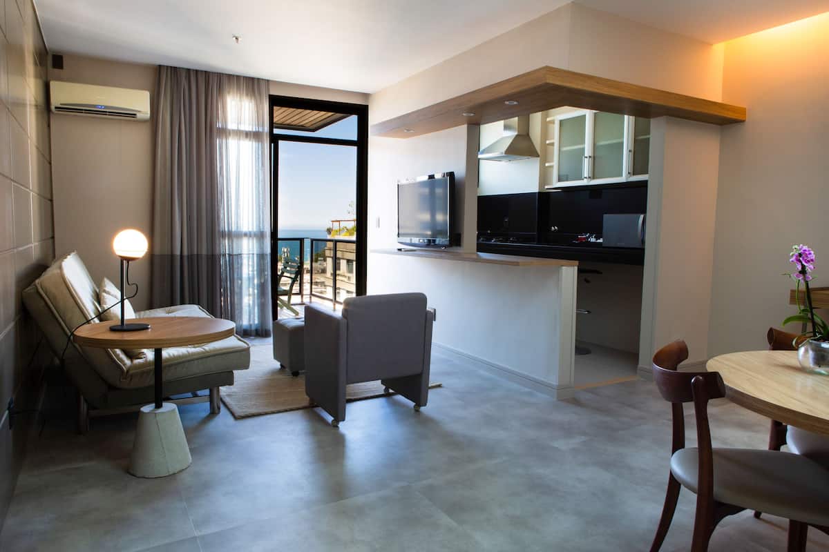 Quarto de hotel moderno com sofá, uma mesinha, TV, área de cozinha e varanda com vista.