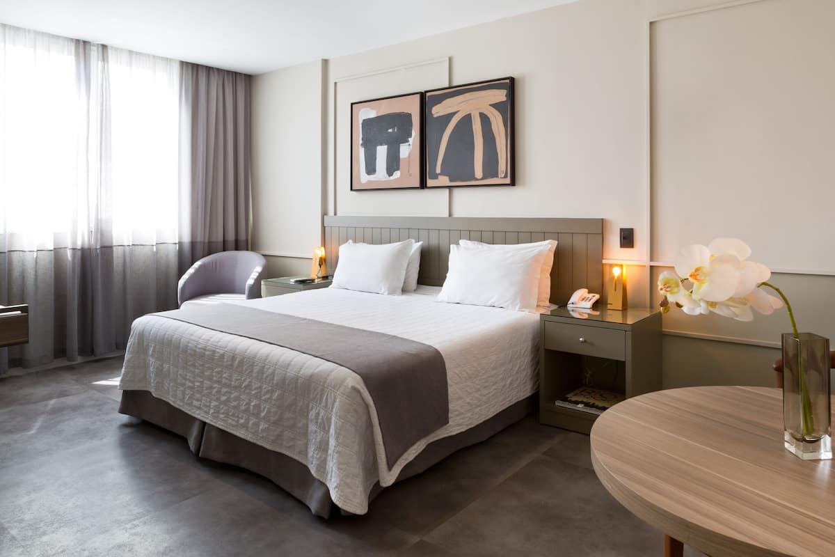 Quarto de hotel com uma cama grande, uma mesa de cabeceira com abajur, uma cadeira e um vaso com flores.