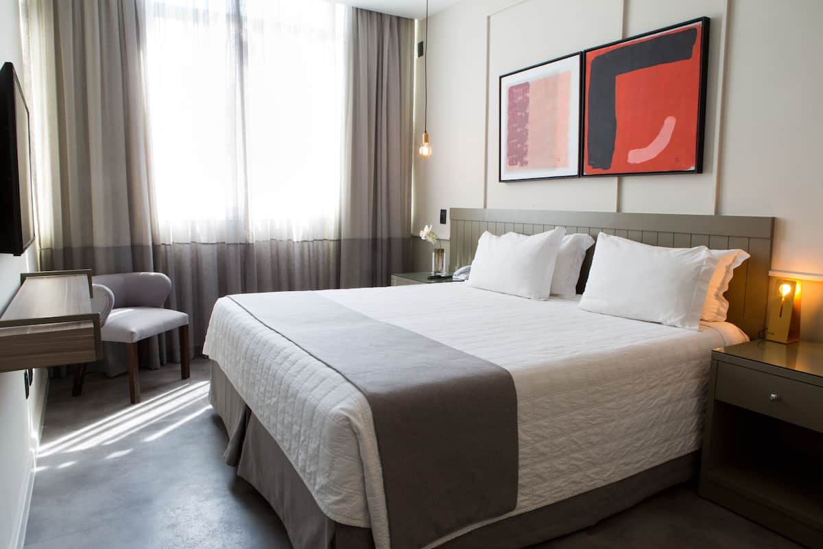 Quarto de hotel com uma cama grande, uma escrivaninha, uma cadeira e uma mesa de cabeceira com abajur.