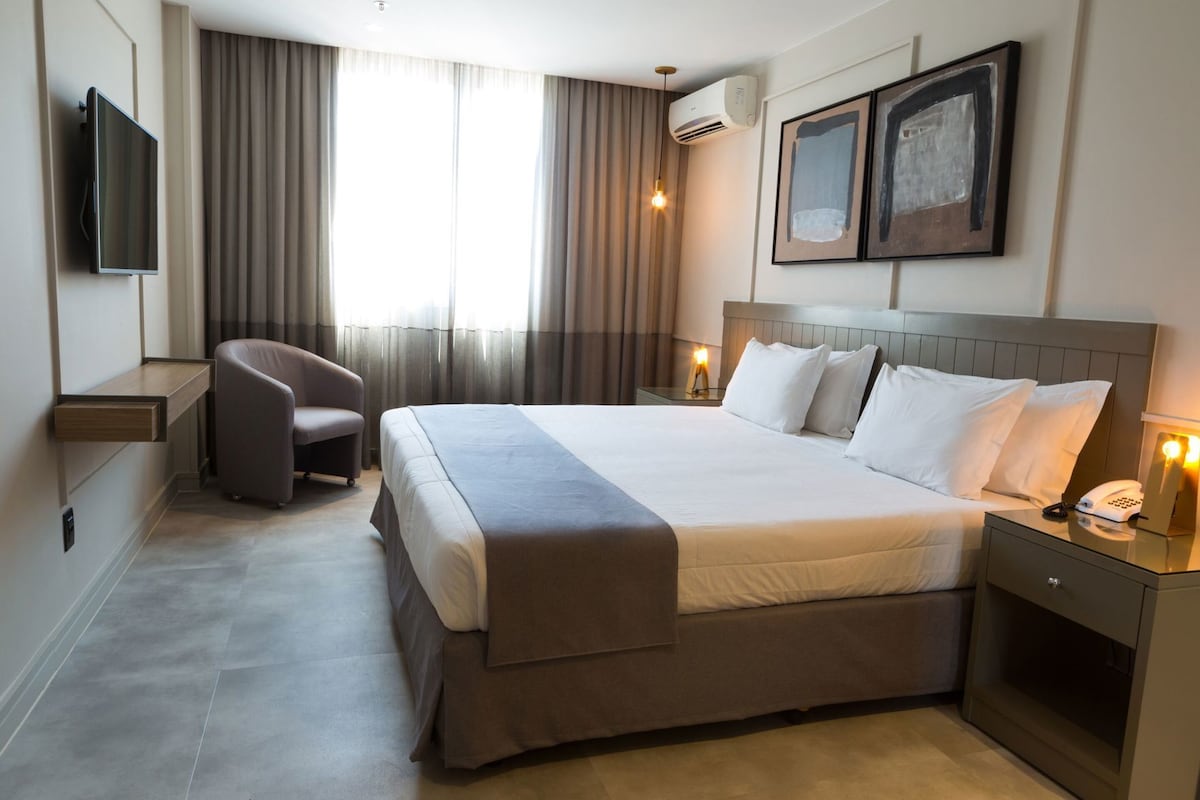 Quarto de hotel com cama, mesa de cabeceira, cadeira, televisão, ar condicionado e obra de arte na parede.