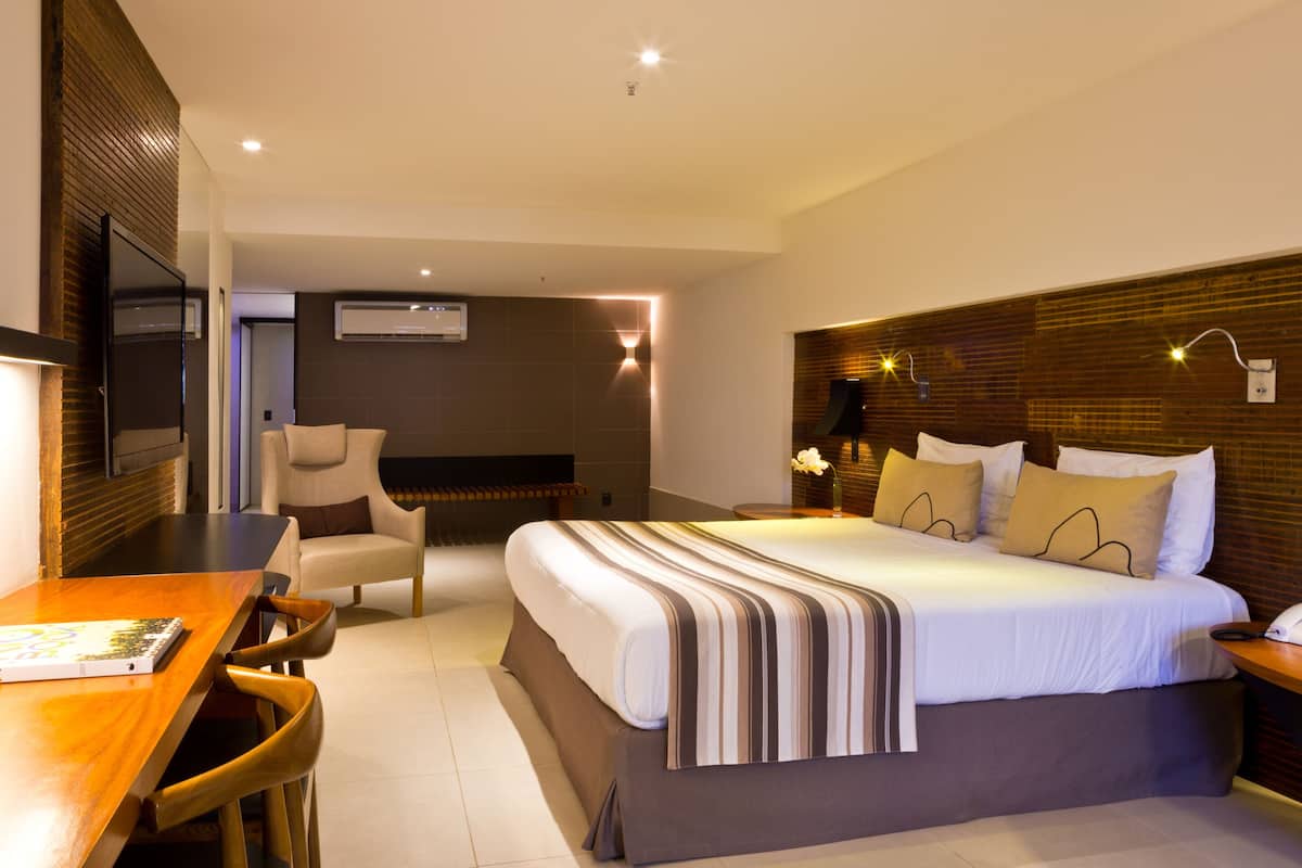 Quarto de hotel moderno com uma cama grande, uma escrivaninha e uma cadeira. Há uma televisão na parede e uma janela.