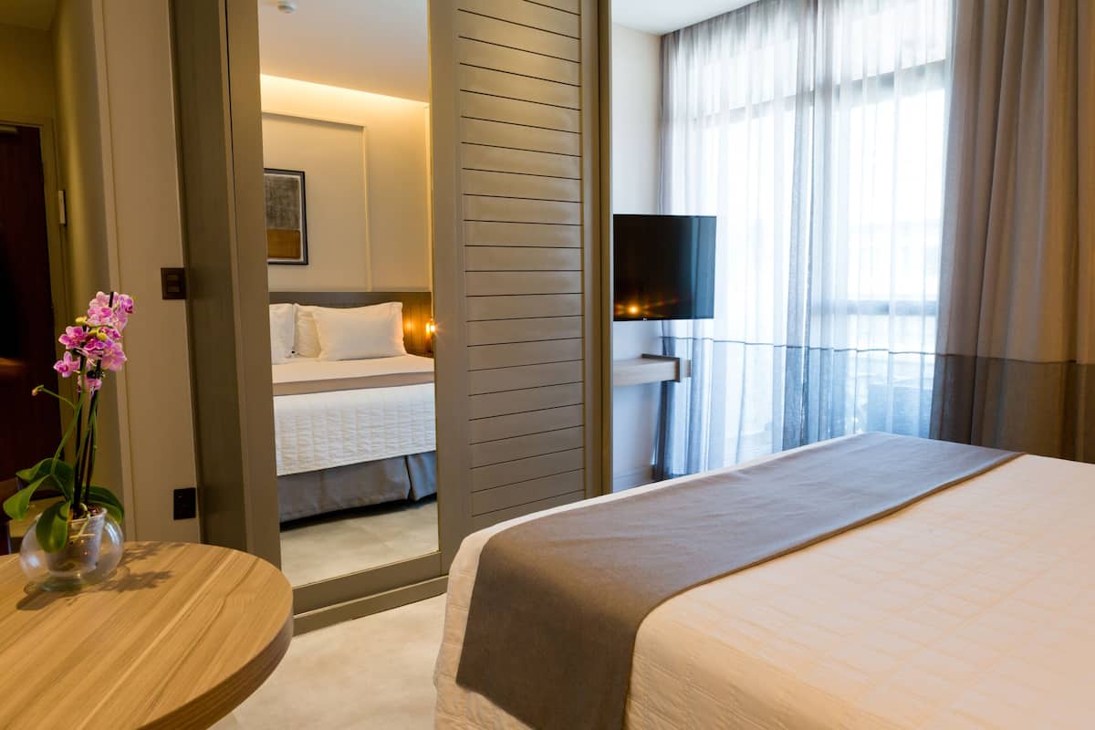 Quarto de hotel com cama, televisão, janela com cortinas e um vaso de flores sobre uma mesa lateral.