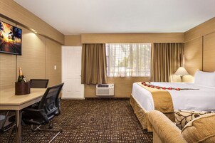 Quarto de hotel com cama, escrivaninha com cadeira, televisão e janela com cortinas.