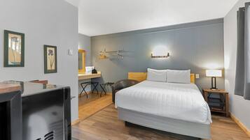 Quarto de hotel com cama, escrivaninha com cadeira, televisão e luminária fixada na parede.