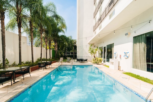 Piscina com espreguiçadeiras e mesas, cercada por palmeiras e um edifício branco.