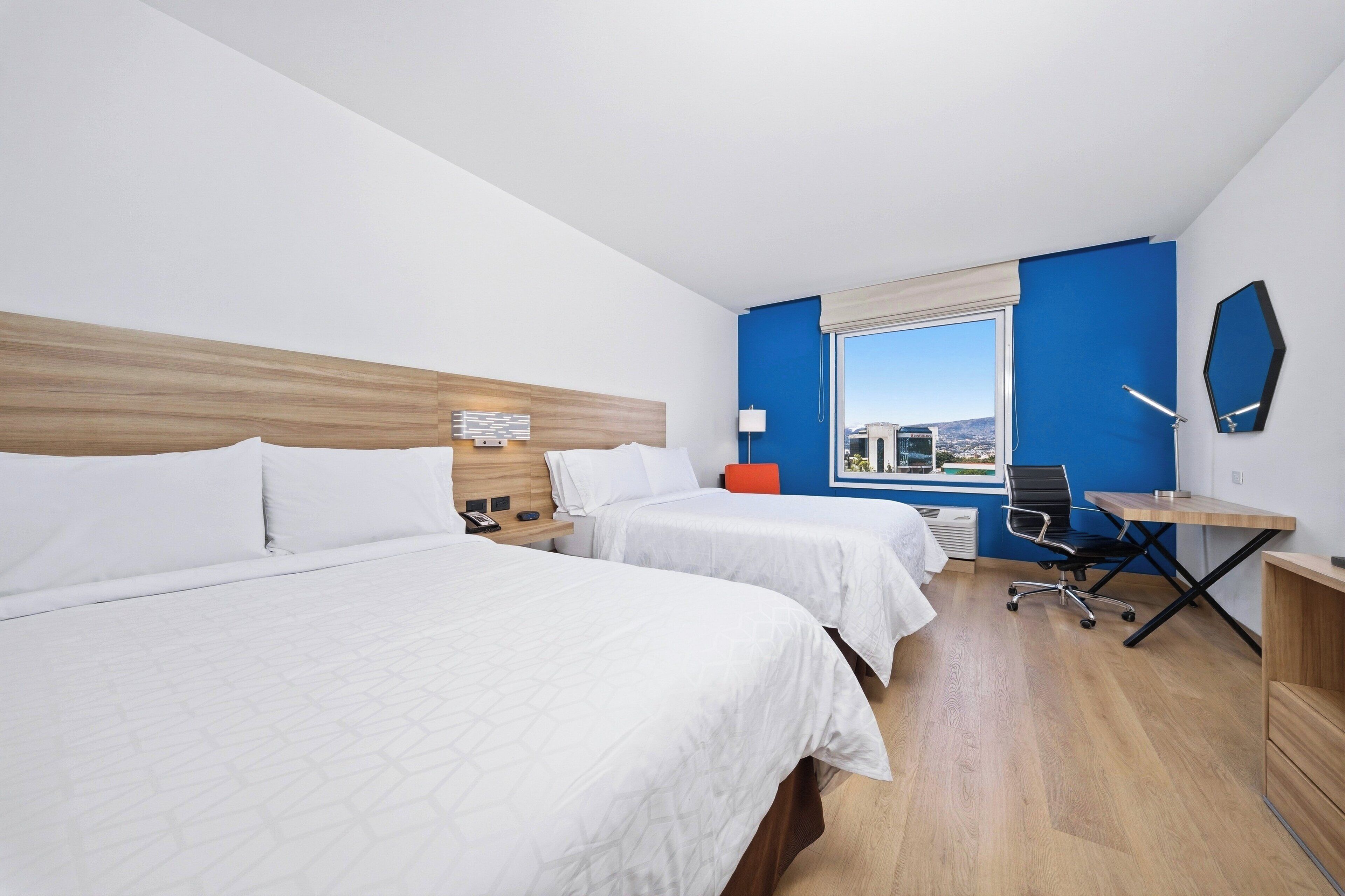 Quarto de hotel com duas camas, uma escrivaninha, uma cadeira, janela com vista e espelho na parede.