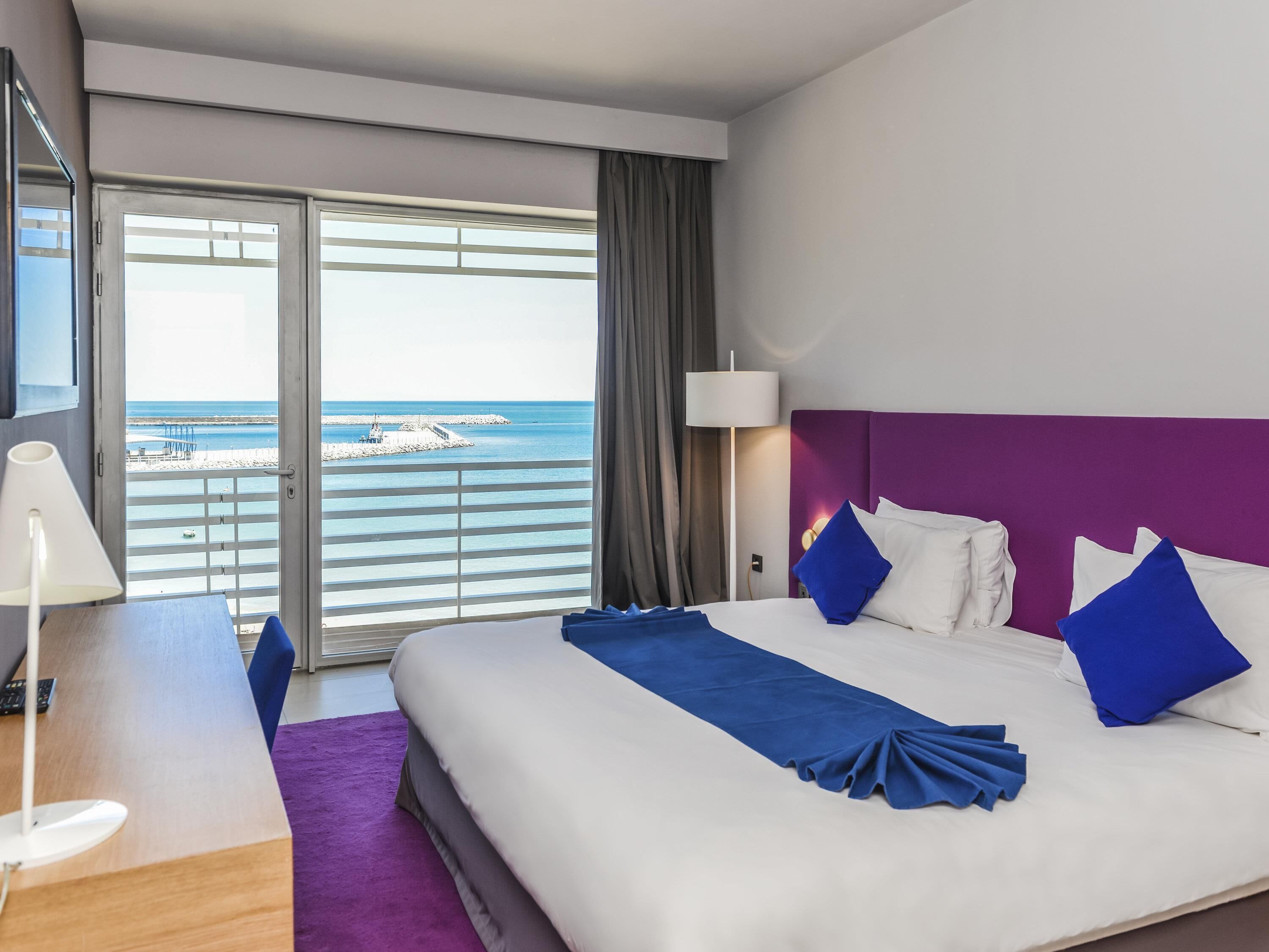 Quarto de hotel com cama, escrivaninha, luminária, vista para o mar e cabeceira roxa.