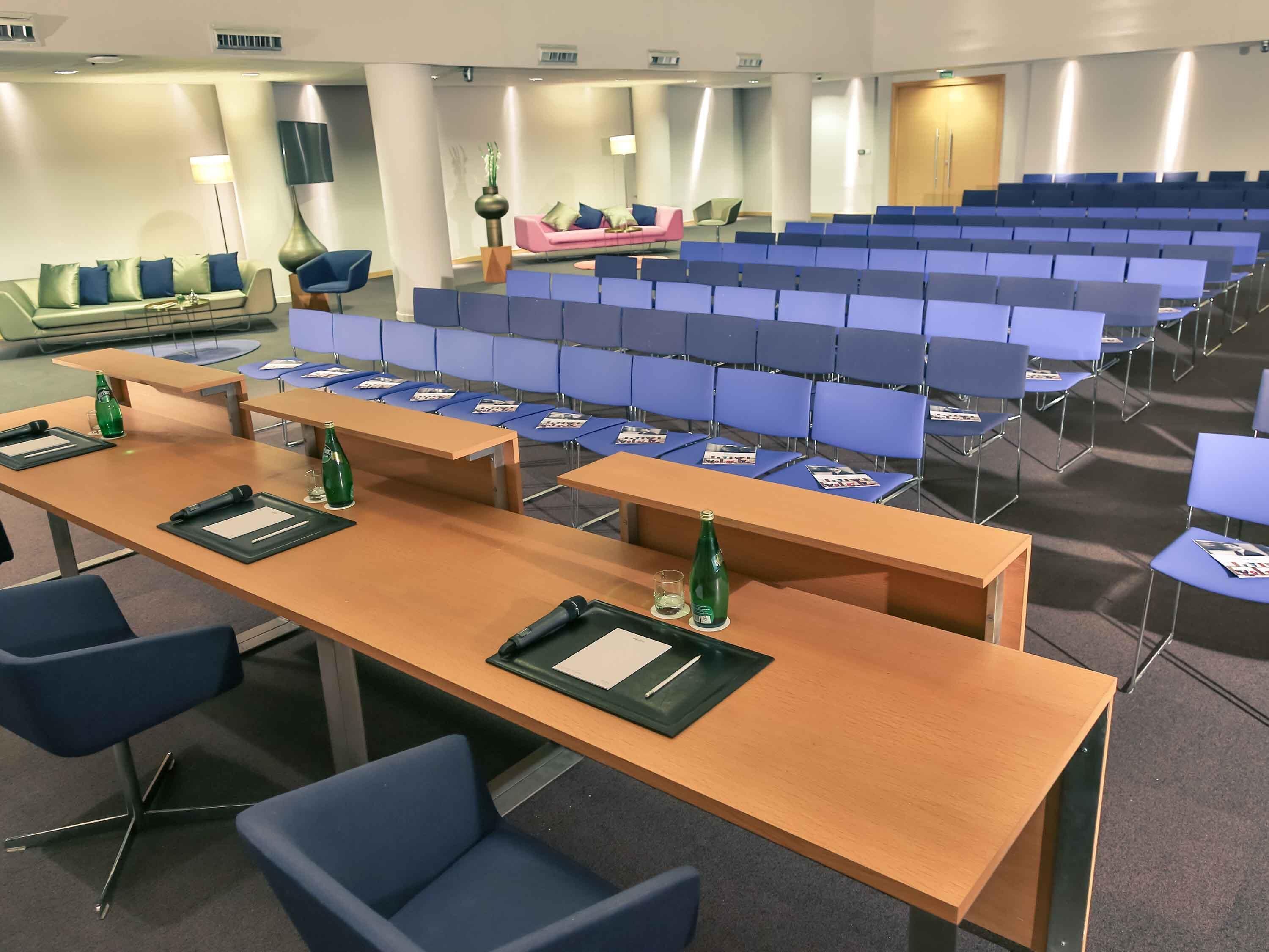 Uma sala de conferências com fileiras de cadeiras azuis, uma mesa comprida com porta-etiquetas e uma área de estar com um sofá rosa e cadeiras.