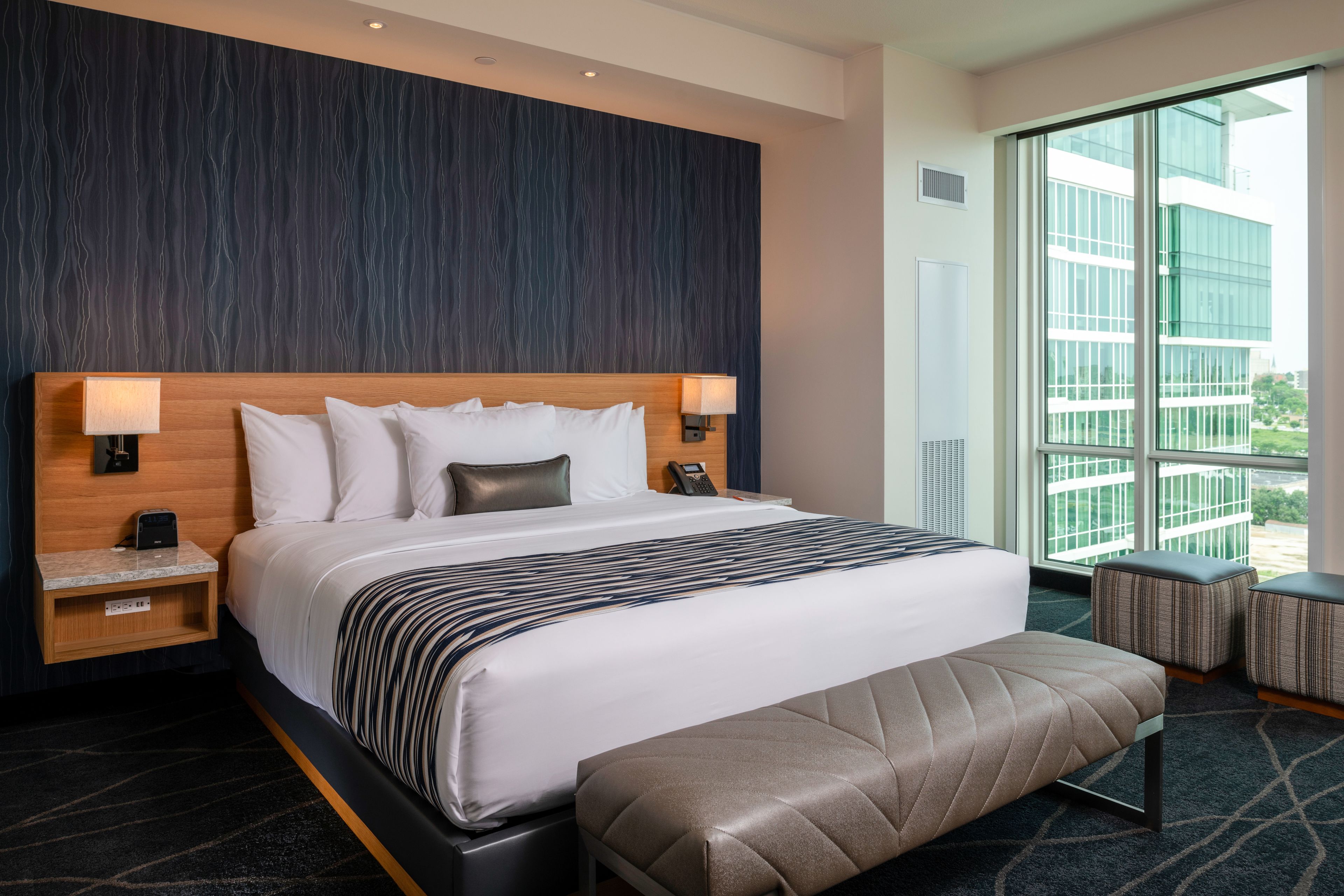Quarto de hotel moderno com uma cama grande, duas mesas de cabeceira com luminárias, um banco e vista para um edifício através de grandes janelas.