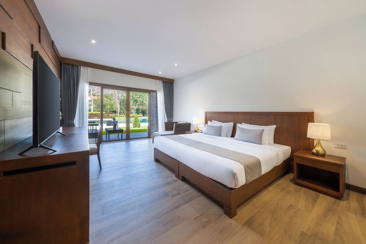 Quarto de hotel com uma cama grande, uma mesa de trabalho com televisão de tela plana, mesa de cabeceira com abajur e porta de vidro corrediça para uma varanda.