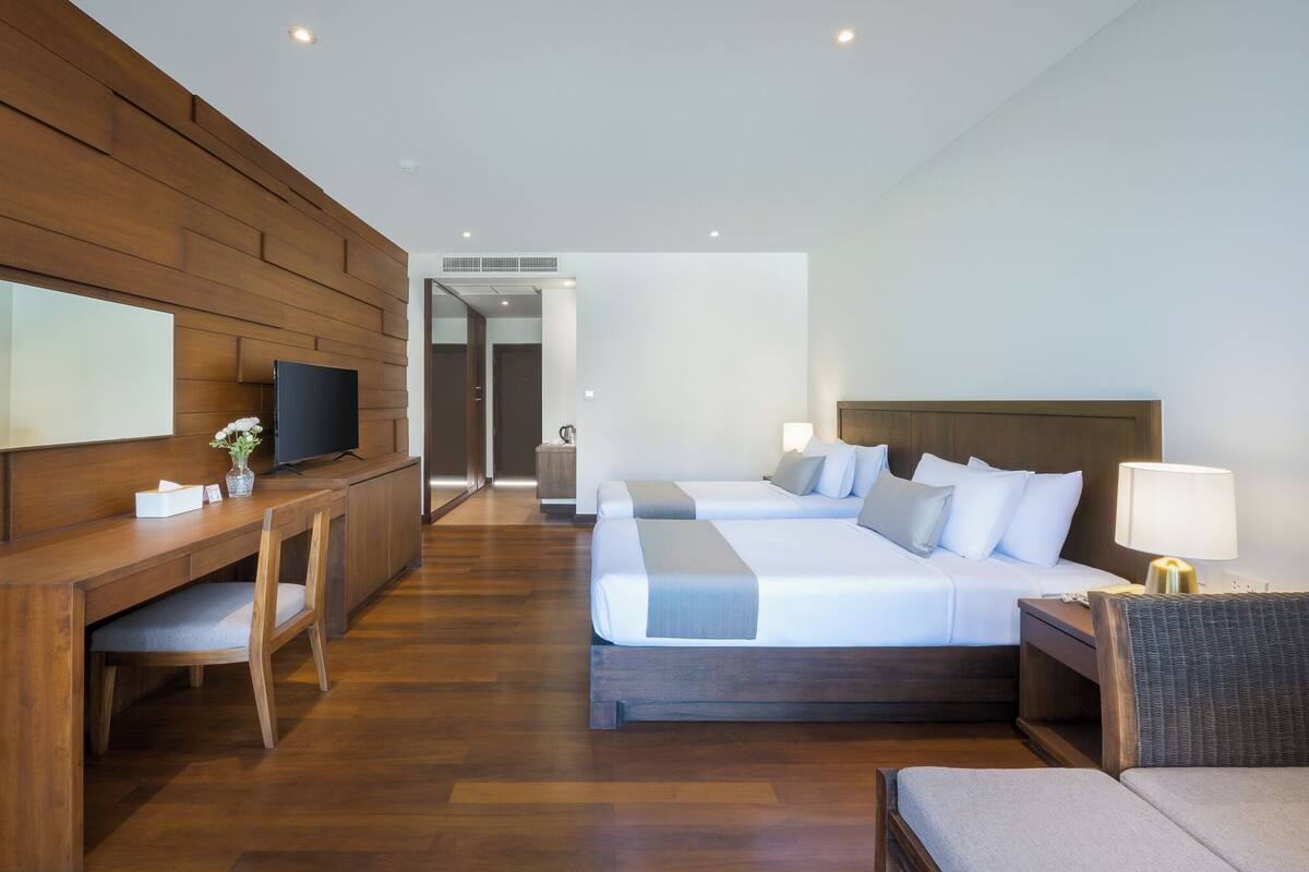 Quarto de hotel com escrivaninha de madeira, duas camas, televisão e mesa de cabeceira com abajur.