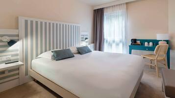 Quarto de hotel com uma cama grande, uma mesa com luminária, uma cadeira e vista para o exterior pela janela.